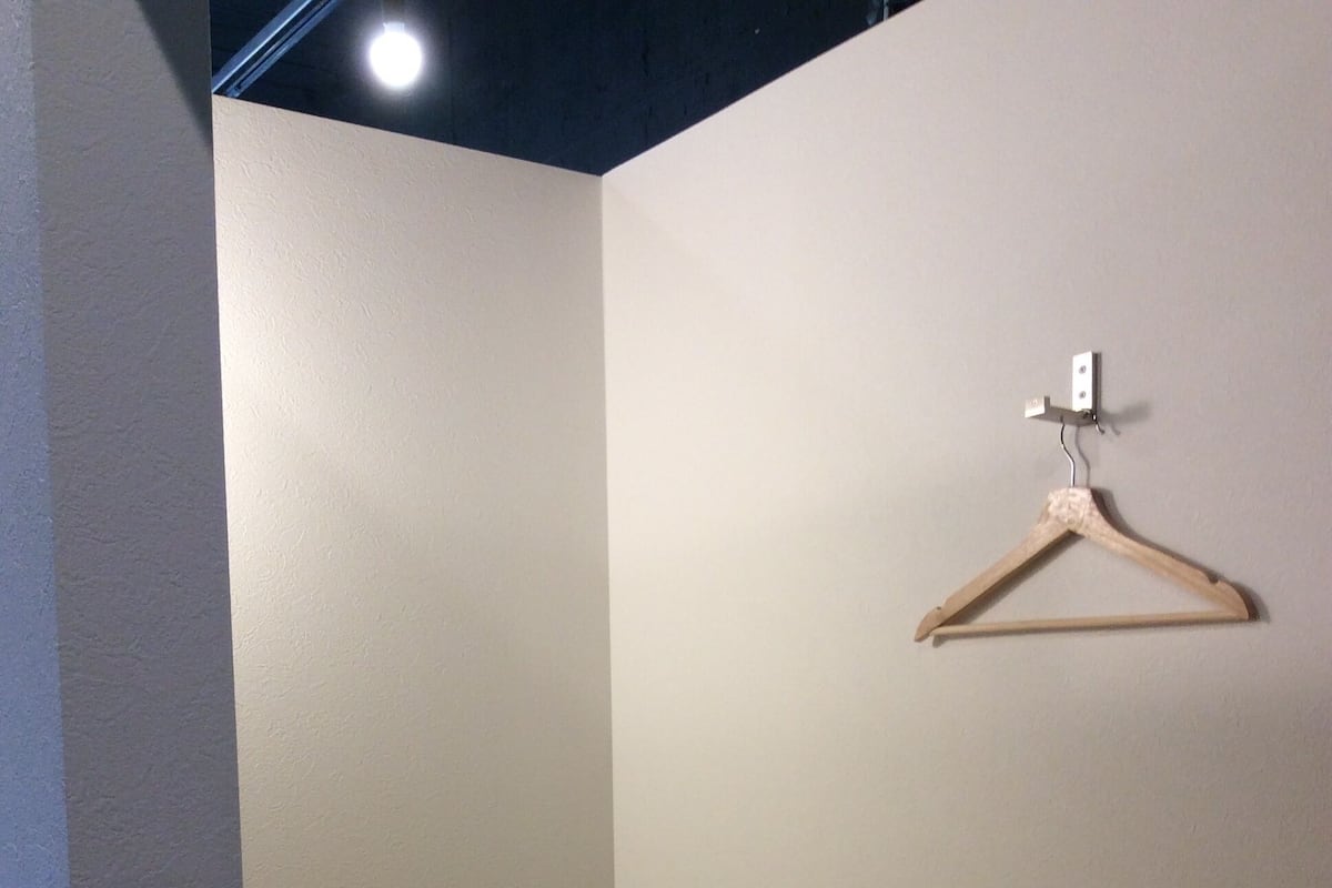 Um quarto pequeno e com pouca luz, com uma cama de solteiro, um cabide de madeira na parede e um lustre.