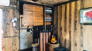 Um banheiro rústico com piso de madeira, vaso sanitário, um banquinho, um violão e uma prateleira com garrafas.
