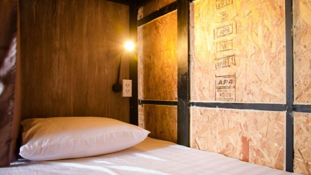 Quarto com parede de madeira, cama com travesseiro branco e luminária fixada na parede.