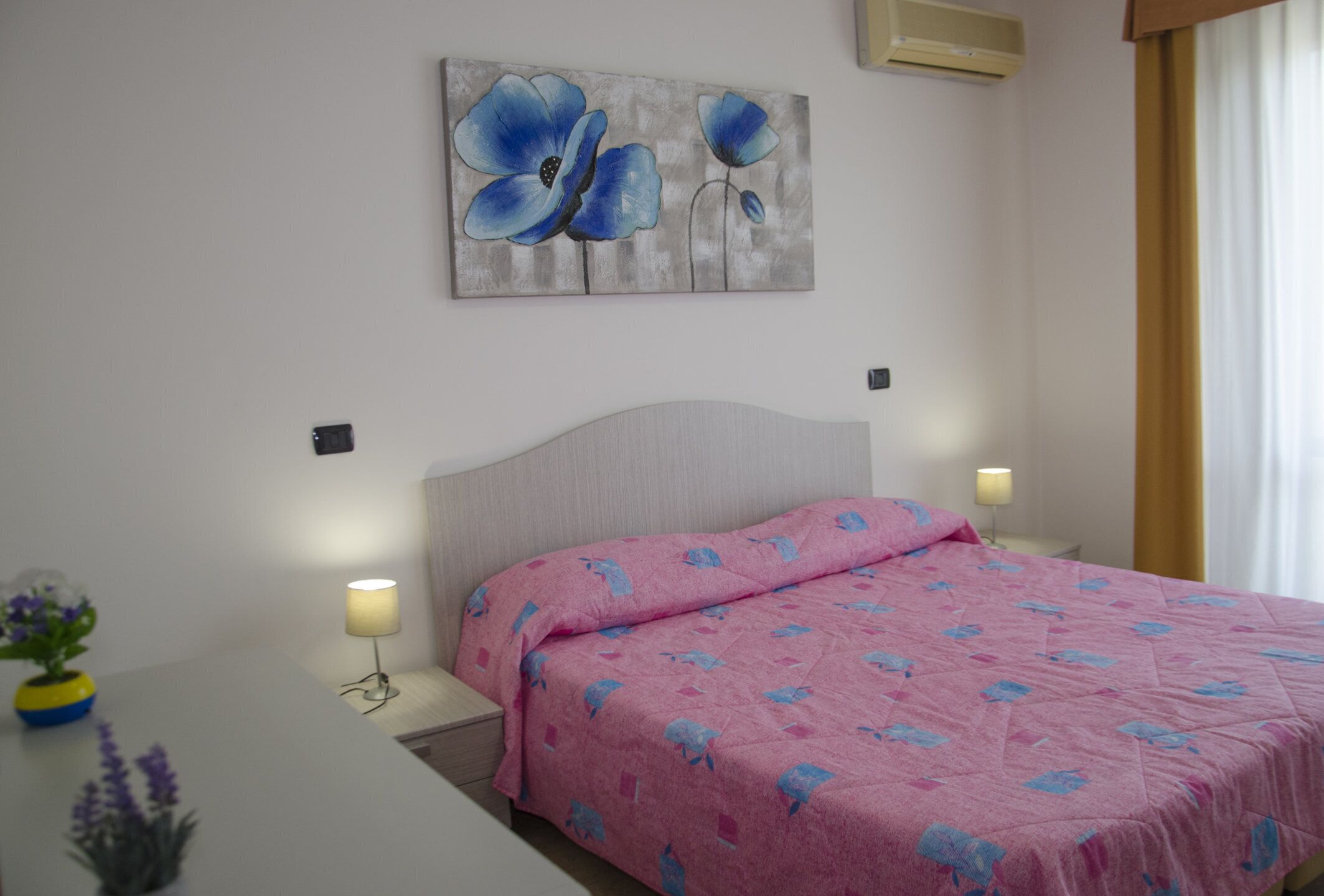 Quarto com cama, abajures, ar condicionado fixado na parede e uma pintura de flores.