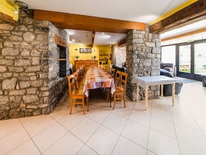 Uma sala de jantar com uma mesa comprida, cadeiras de madeira, paredes de pedra e vista através de uma porta de vidro.