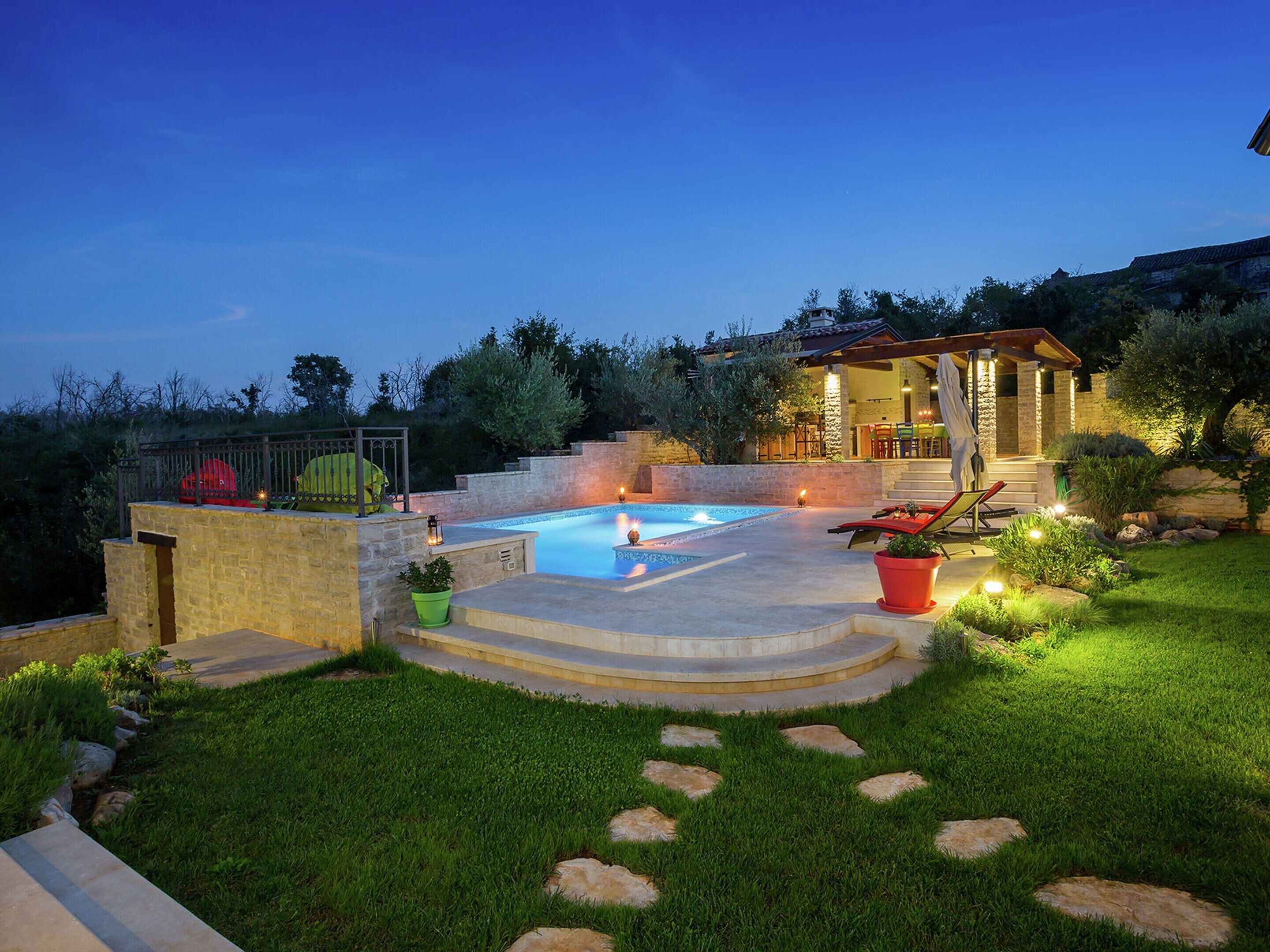 Uma área de piscina ao ar livre bem iluminada, com deck de pedra, uma espreguiçadeira e um pequeno pátio com mesa e cadeiras.