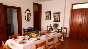 Uma sala de jantar com uma mesa posta para seis lugares, cadeiras, uma cozinha ao fundo e duas pinturas emolduradas na parede.