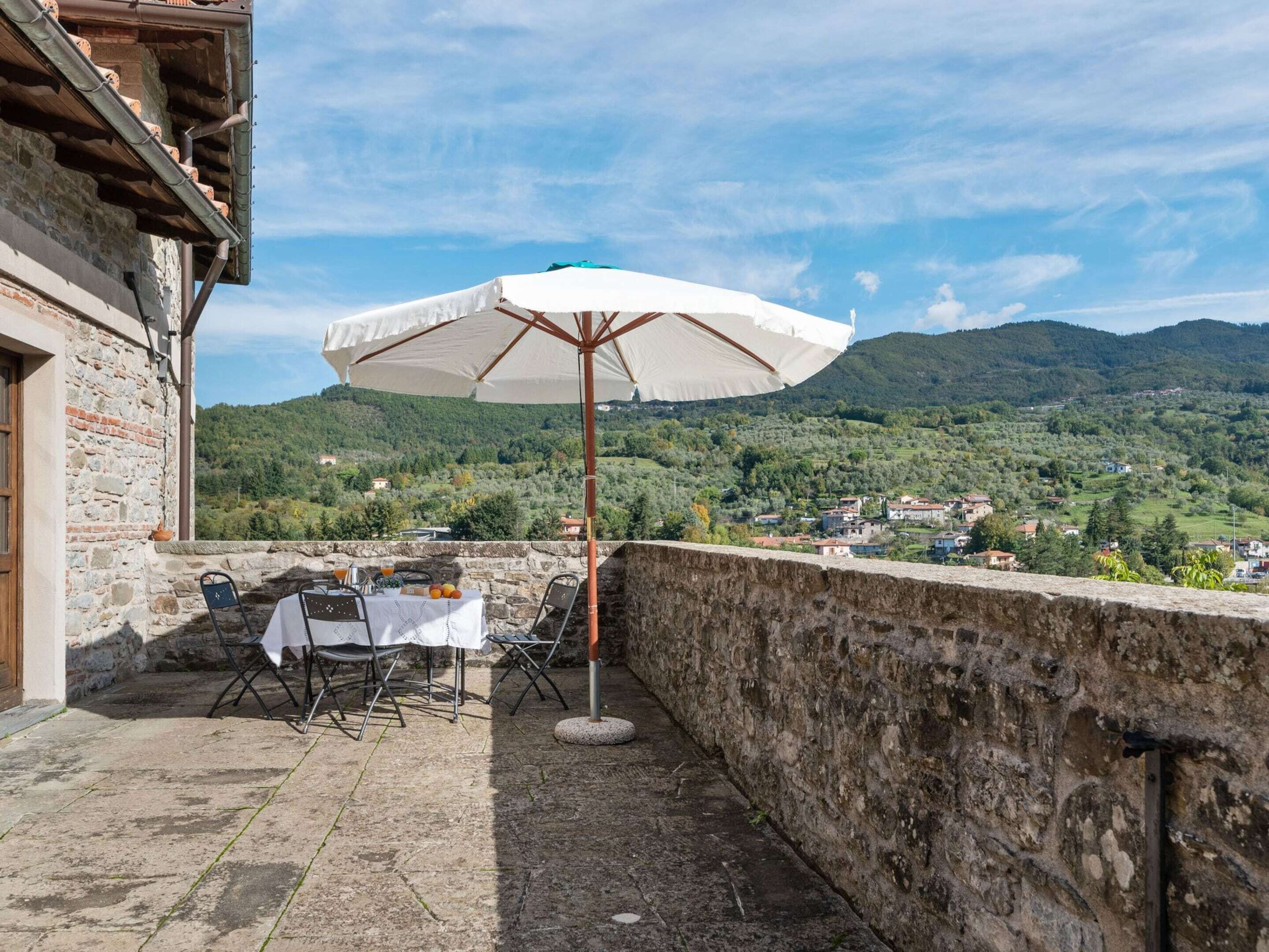 Um terraço de pedra com mesa e cadeiras, um guarda-sol e vista para uma aldeia no morro.
