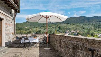 Um terraço de pedra com mesa e cadeiras, um guarda-sol e vista para uma aldeia no morro.
