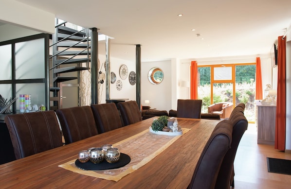 Uma sala de jantar moderna com mesa de madeira, cadeiras de couro, escada em espiral e vista através de grandes janelas.