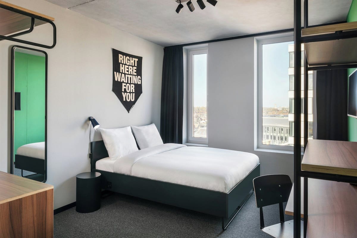 Quarto de hotel moderno com cama, escrivaninha, cadeira e uma janela grande com vista para a cidade.