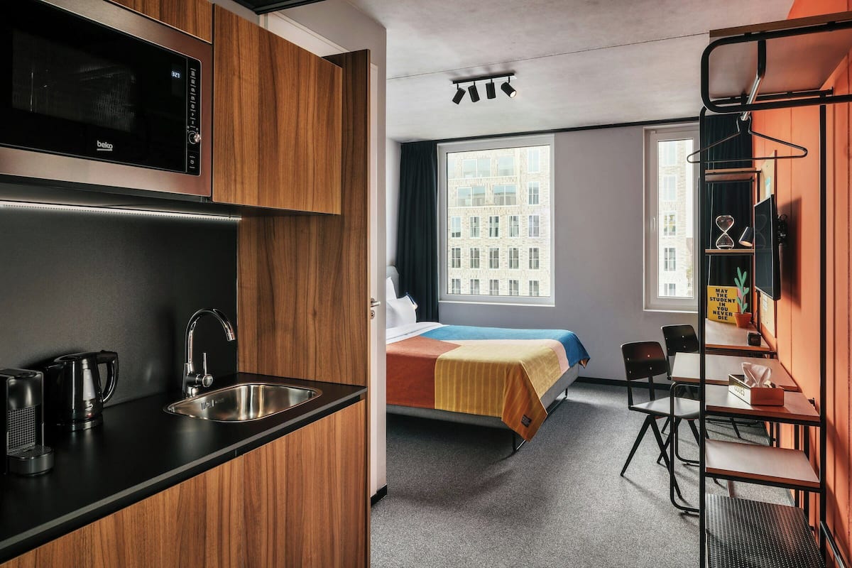 Quarto de hotel compacto com cozinha compacta, uma cama com colcha colorida, uma escrivaninha com cadeira e vista para um edifício pela janela.