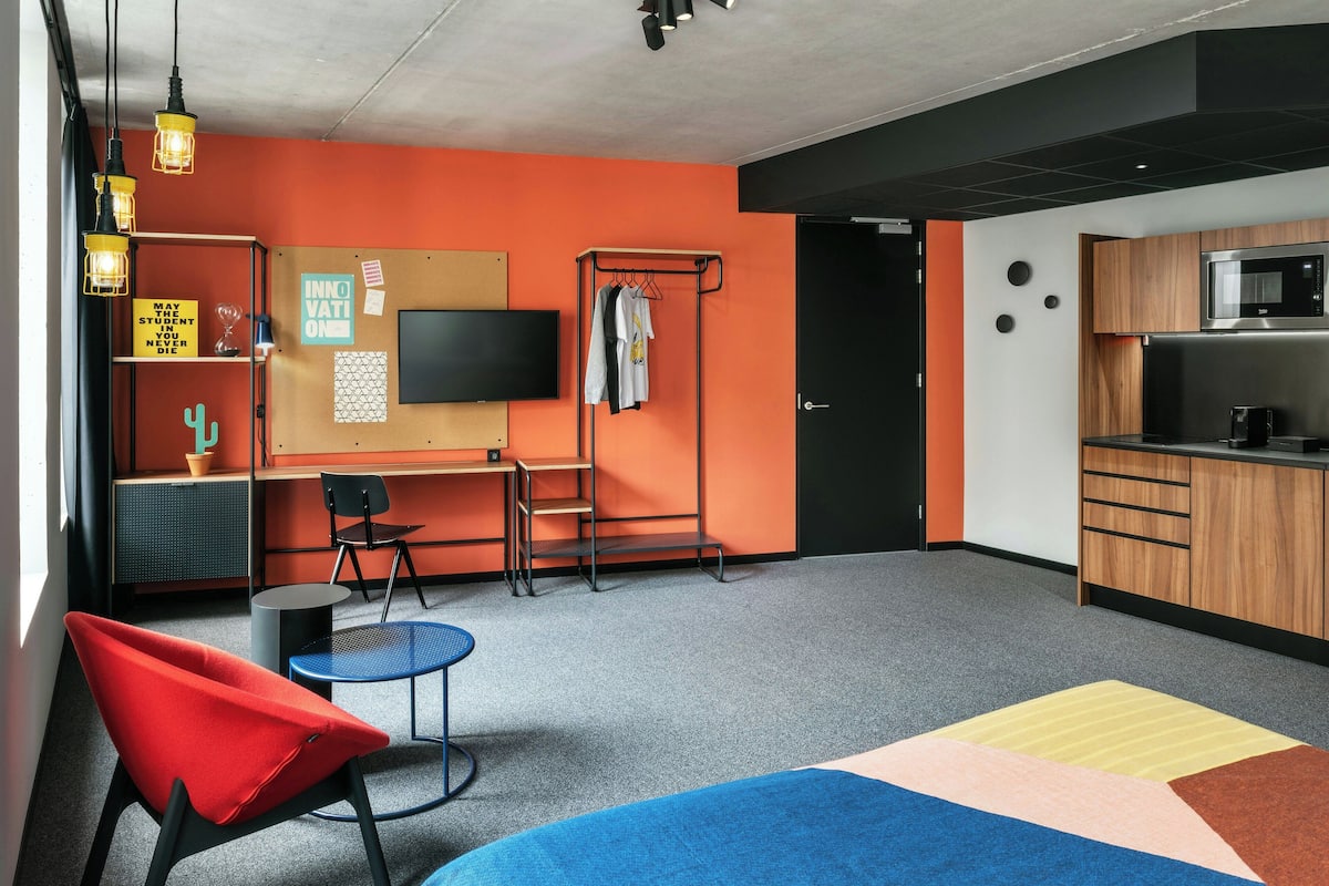 Quarto moderno com escrivaninha, cadeira e televisão. Possui cozinha compacta com armários e micro-ondas.