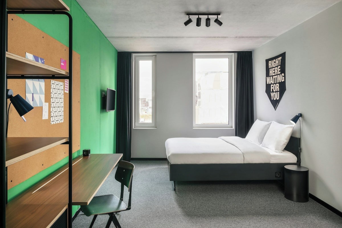 Quarto de hotel moderno com cama, escrivaninha, cadeira e um painel de cortiça com anotações.