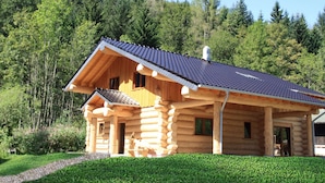 Uma cabana de toras com telhado inclinado e um pequeno alpendre, rodeada por um jardim bem cuidado e uma floresta densa.