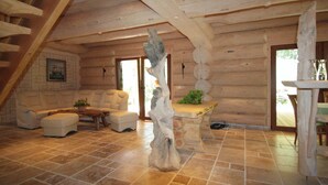 Interior de cabana de tronco com piso de ladrilho de pedra, escada de madeira, sofá e mesa com uma planta.
