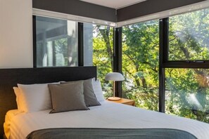 Quarto de hotel moderno com uma cama grande, uma mesinha de cabeceira e uma luminária. O quarto possui janelas amplas com vista para árvores.