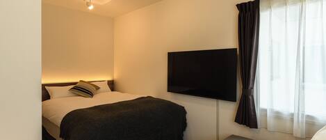 Quarto de hotel com cama, TV de tela plana na parede, janela com cortinas e um pequeno espaço para sentar.