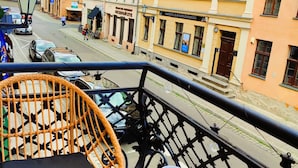 Varanda com cadeiras de vime, uma mesa com planta em vaso e vista para a rua com edifícios e carros estacionados.