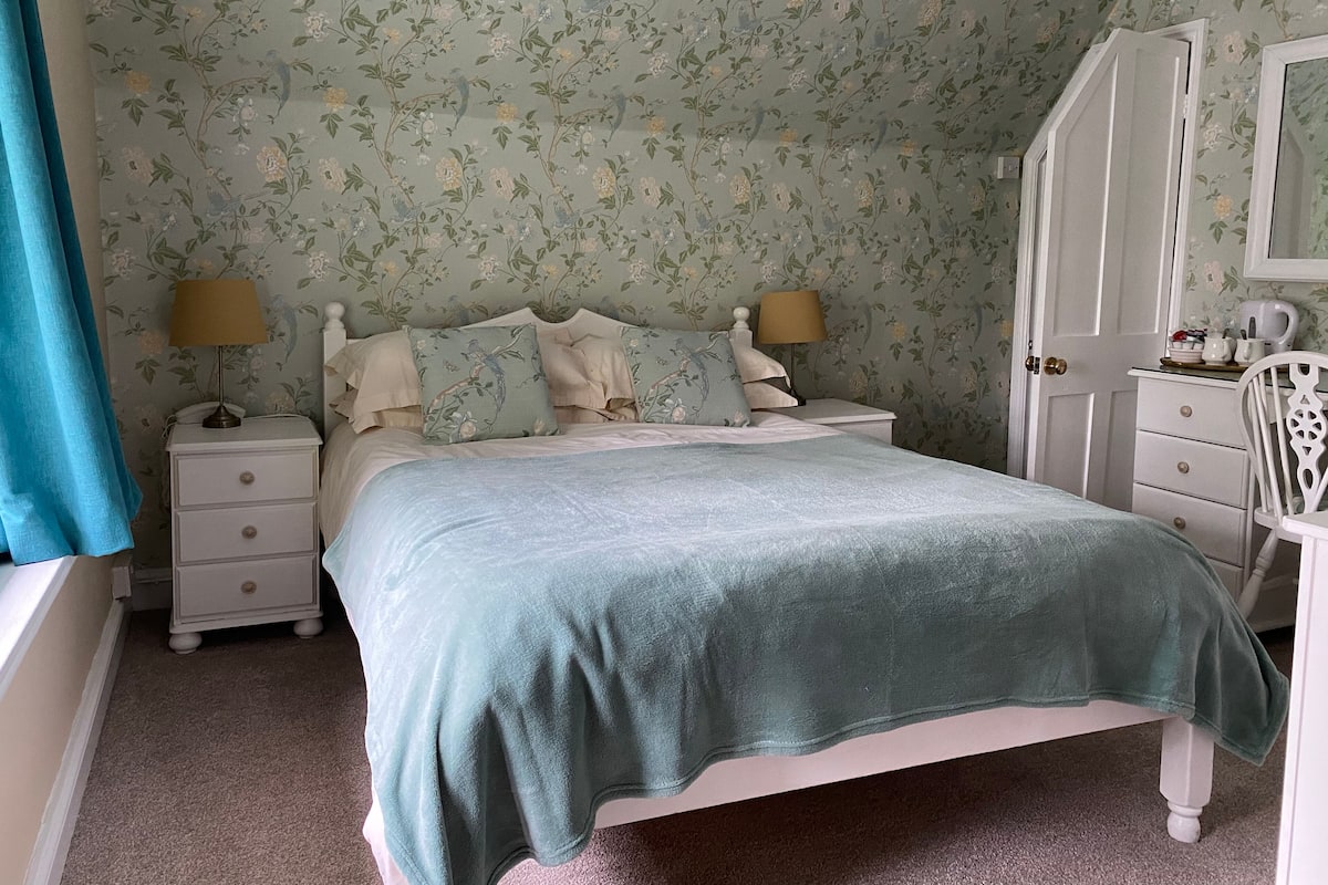 Quarto com papel de parede floral, cama com manta verde, duas luminárias de cabeceira, cômoda branca e uma cadeira.