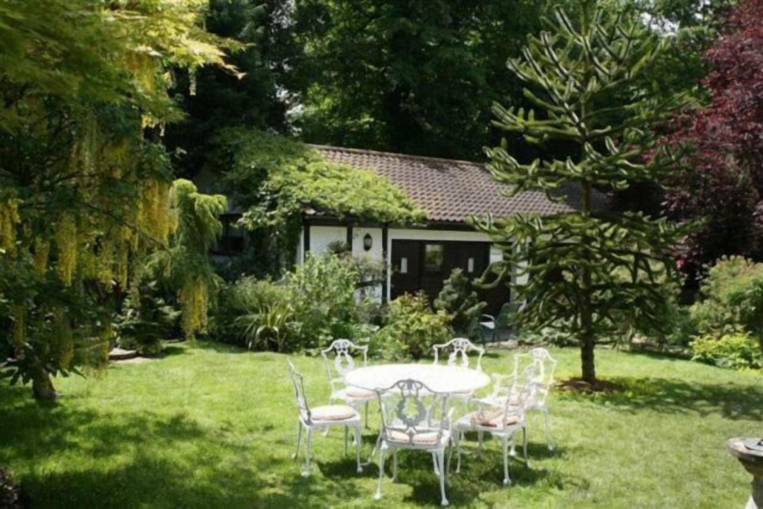 Um jardim com mesa e cadeiras brancas, cercado por vegetação e um edifício com telhado em telhas.