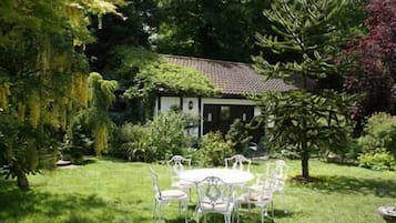 Um jardim com mesa e cadeiras brancas, cercado por vegetação e um edifício com telhado em telhas.