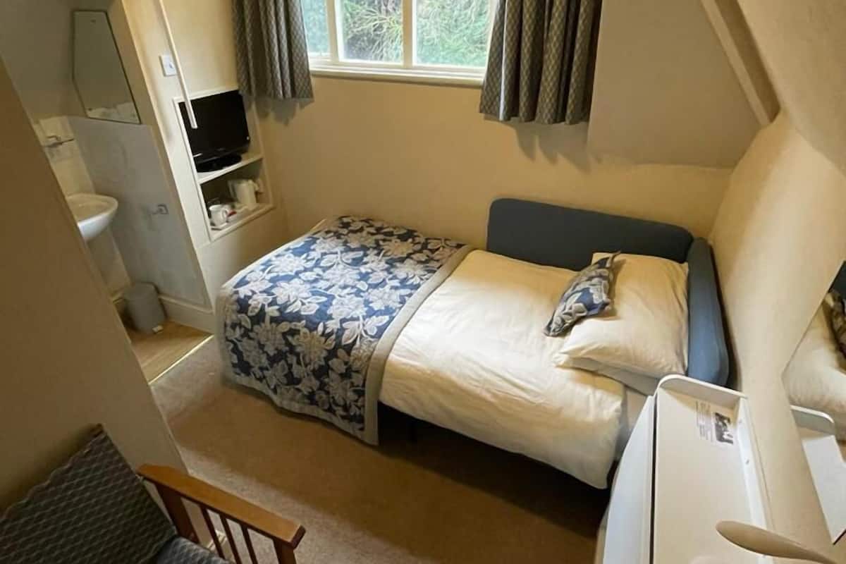 Quarto com cama, escrivaninha, cadeira, televisão e janela com cortinas.