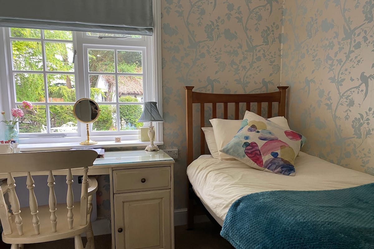 Quarto com cama de madeira, escrivaninha com cadeira, janela com vista para vegetação e papel de parede floral.