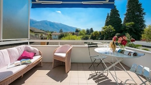 Uma varanda com sofá, cadeira, mesa e flores; vista para as montanhas ao fundo.