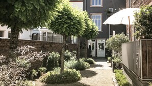 Uma passagem estreita que leva a uma casa com fachada escura, porta branca e um pequeno jardim com diversas plantas.