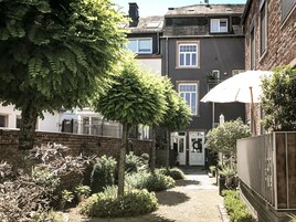 Uma passagem estreita que leva a uma casa com fachada escura, porta branca e um pequeno jardim com diversas plantas.