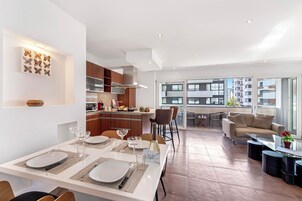 Uma sala de estar moderna com área de refeições, janelas amplas e cozinha ao fundo.