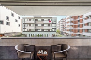 Uma varanda com duas cadeiras de vime e uma pequena mesa posta para dois, com vista para um prédio moderno com várias varandas e janelas.