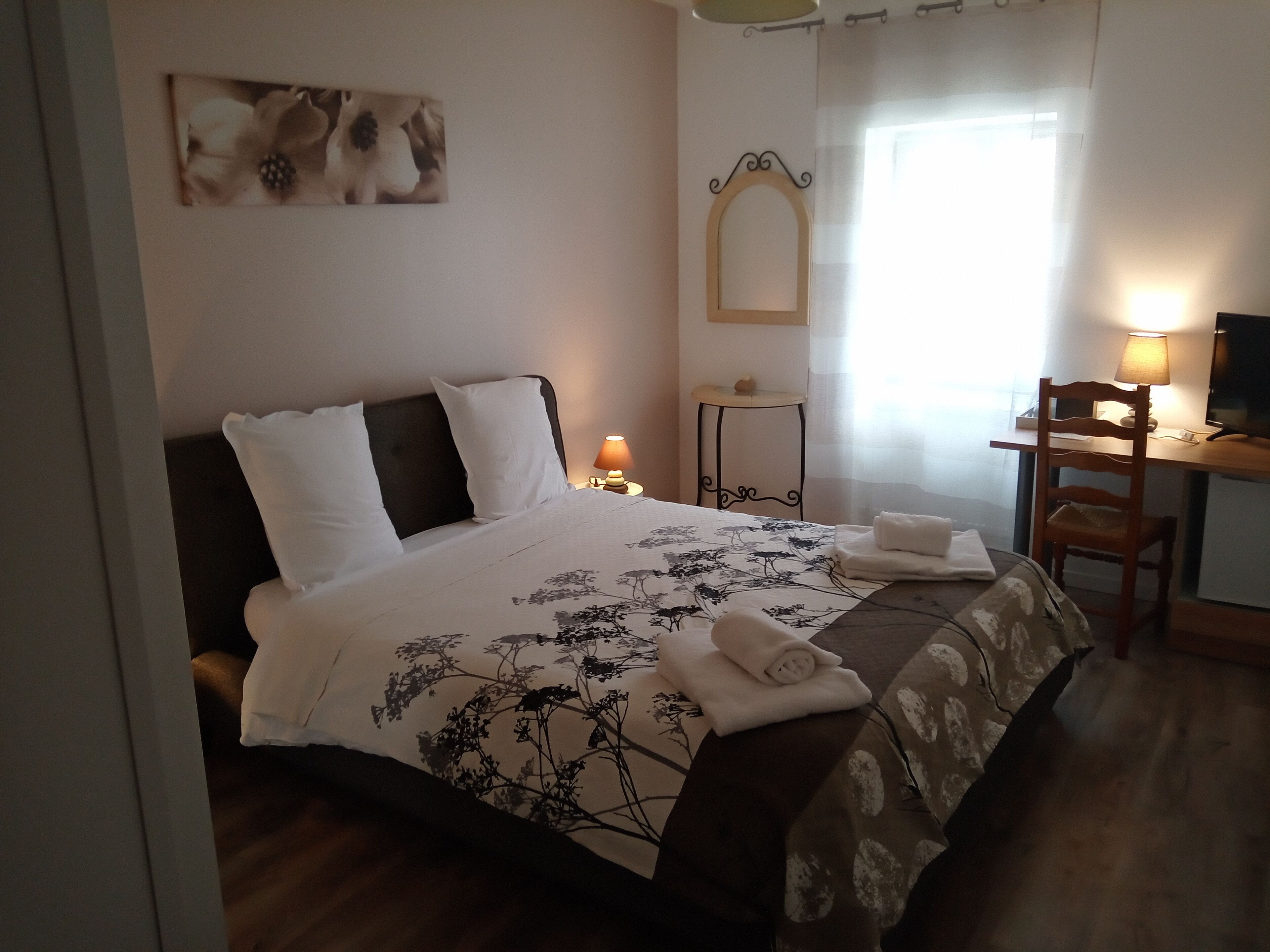 Uma cama bem arrumada com um cobreleito floral, uma mesa de cabeceira com abajur, uma cadeira e uma escrivaninha com um computador.