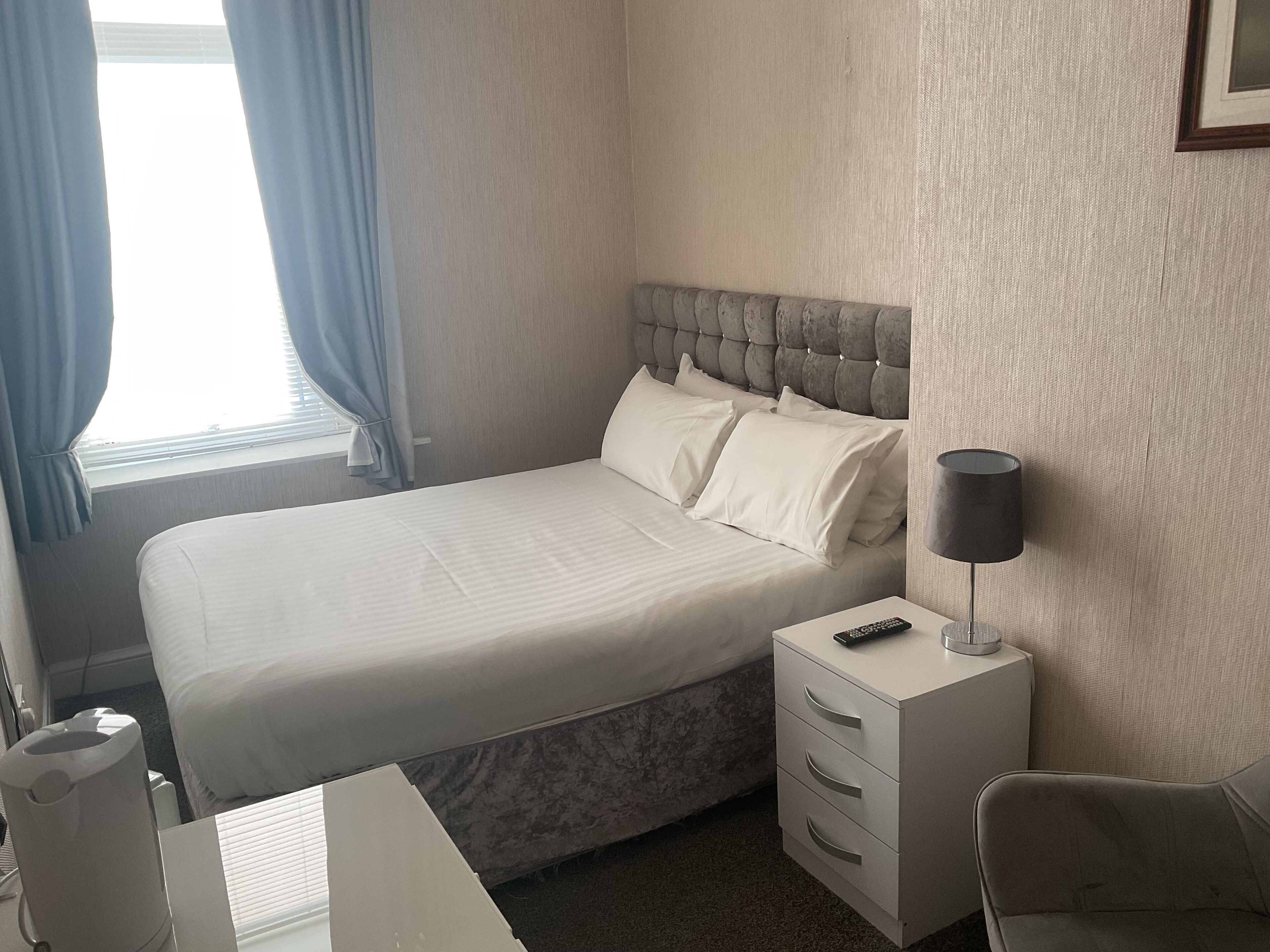 Quarto de hotel bem arrumado, com cama, mesa de cabeceira, luminária e janela com cortinas.