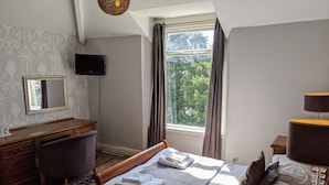 Quarto de hotel com cama, escrivaninha, cadeira, televisão e janela com cortinas.