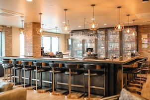 Uma área de bar moderna com ilha central, vários bancos altos, iluminação pendente e um painel de parede de tijolos.
