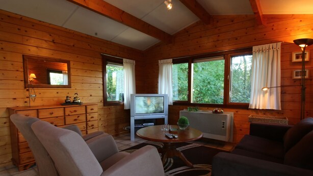 Interior acolhedor de uma cabana, com paredes de madeira, sala de jantar com mesa e cadeiras, televisão e uma grande janela com cortinas.