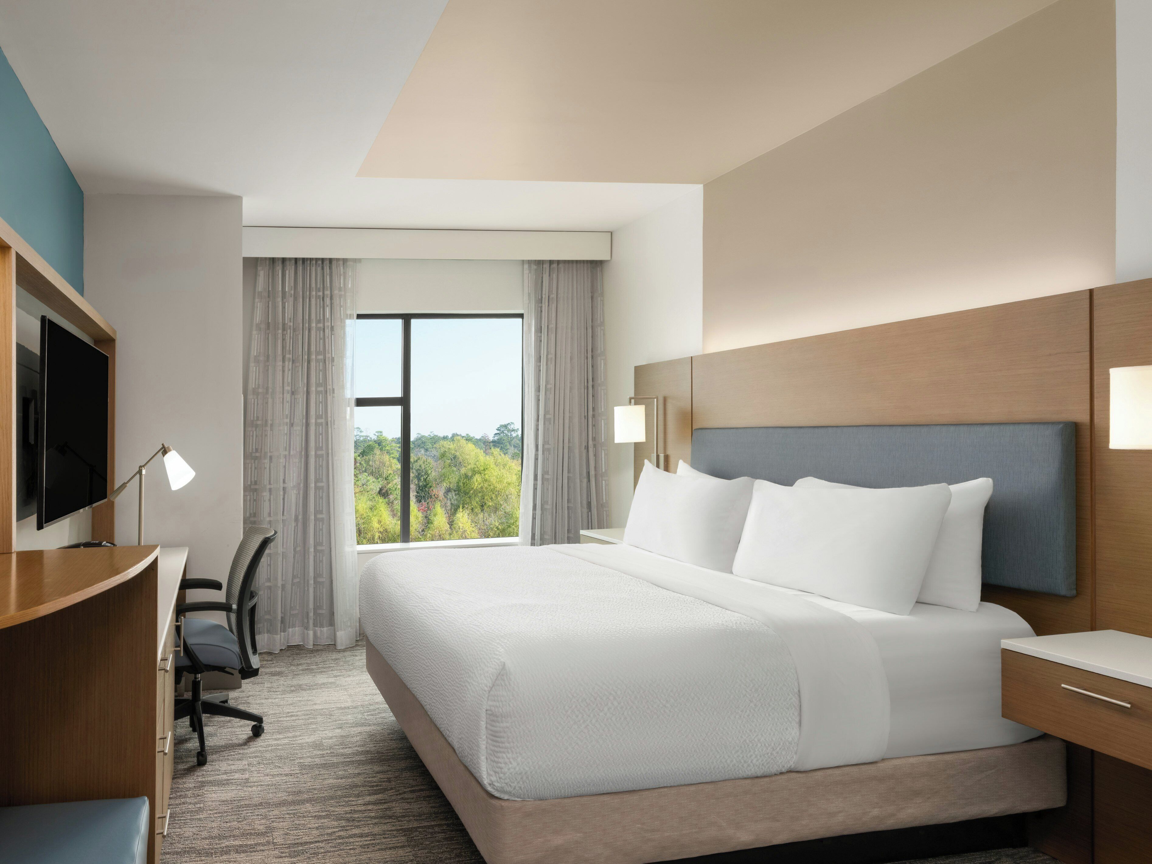 Quarto de hotel com uma cama grande, uma escrivaninha, uma cadeira, televisão e uma janela com cortinas.