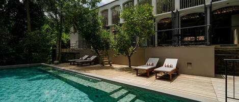 Piscina com espreguiçadeiras, rodeada por árvores e um edifício com grandes janelas.
