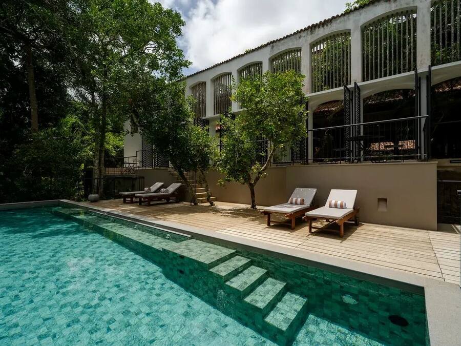 Piscina com espreguiçadeiras, rodeada por árvores e um edifício com grandes janelas.