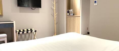 Quarto de hotel com uma cama, televisão na parede, porta-roupas, armário de madeira e uma pequena prateleira com uma chaleira.