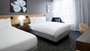 Quarto de hotel com duas camas, uma escrivaninha, uma cadeira, um abajur e uma janela grande com cortinas.