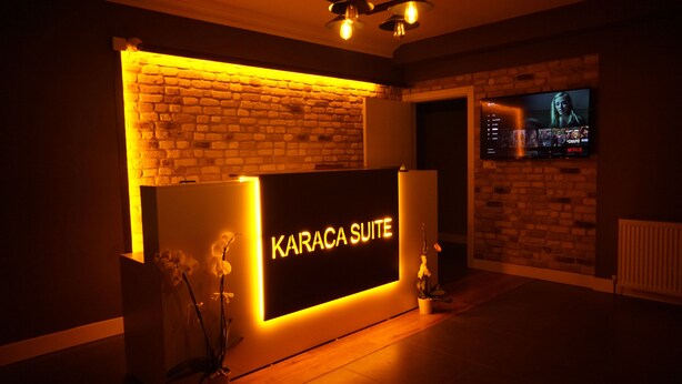 Uma receção com um letreiro de néon a indicar "KARACA SUITE" num hall moderno com parede de tijolo e televisão de ecrã plano fixada na parede.