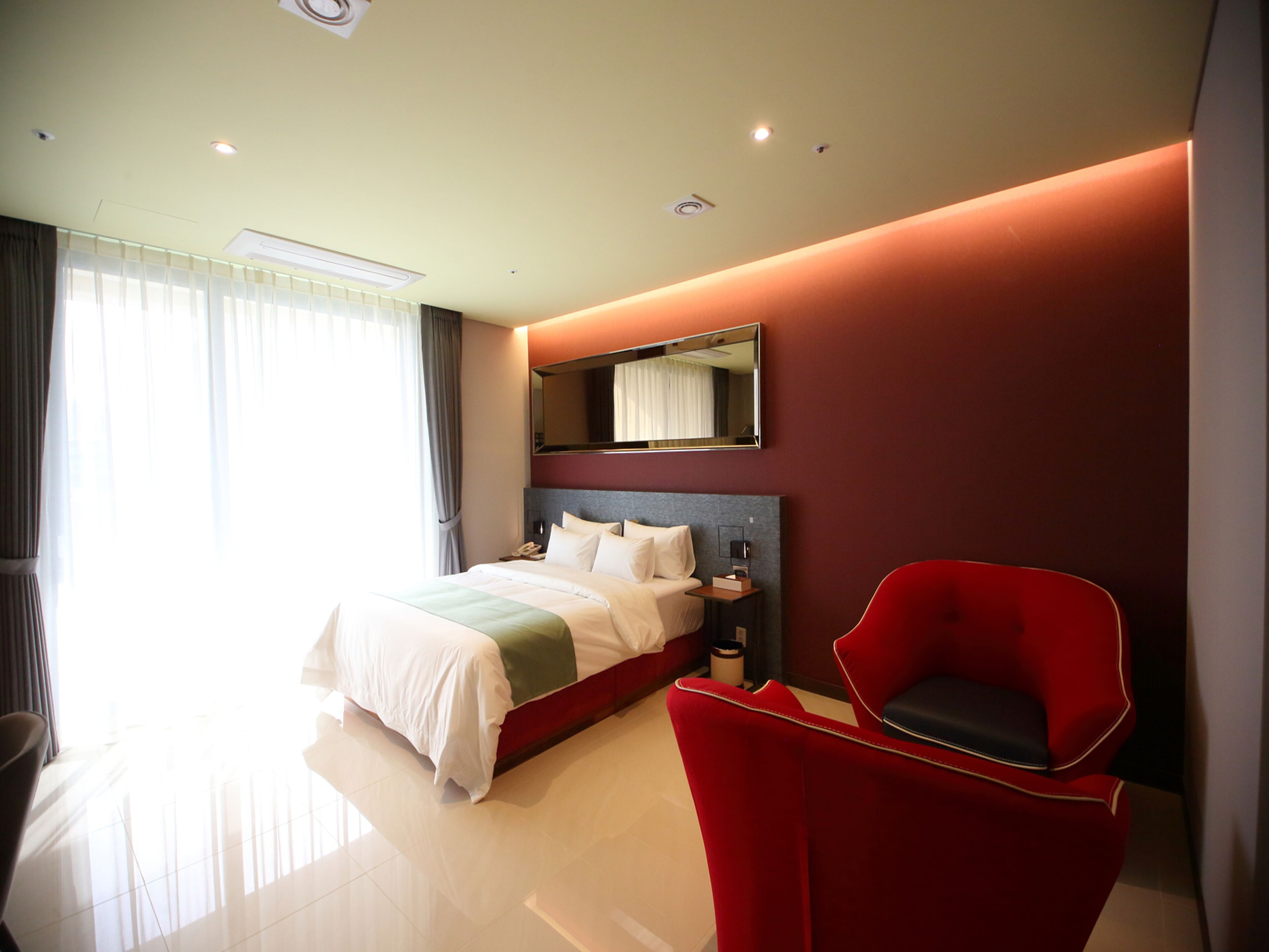 Quarto de hotel moderno com uma cama grande, uma poltrona vermelha, uma mesinha de cabeceira e um espelho na parede.