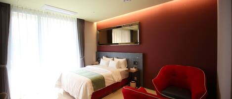 Quarto de hotel moderno com uma cama grande, uma poltrona vermelha, uma mesinha de cabeceira e um espelho na parede.
