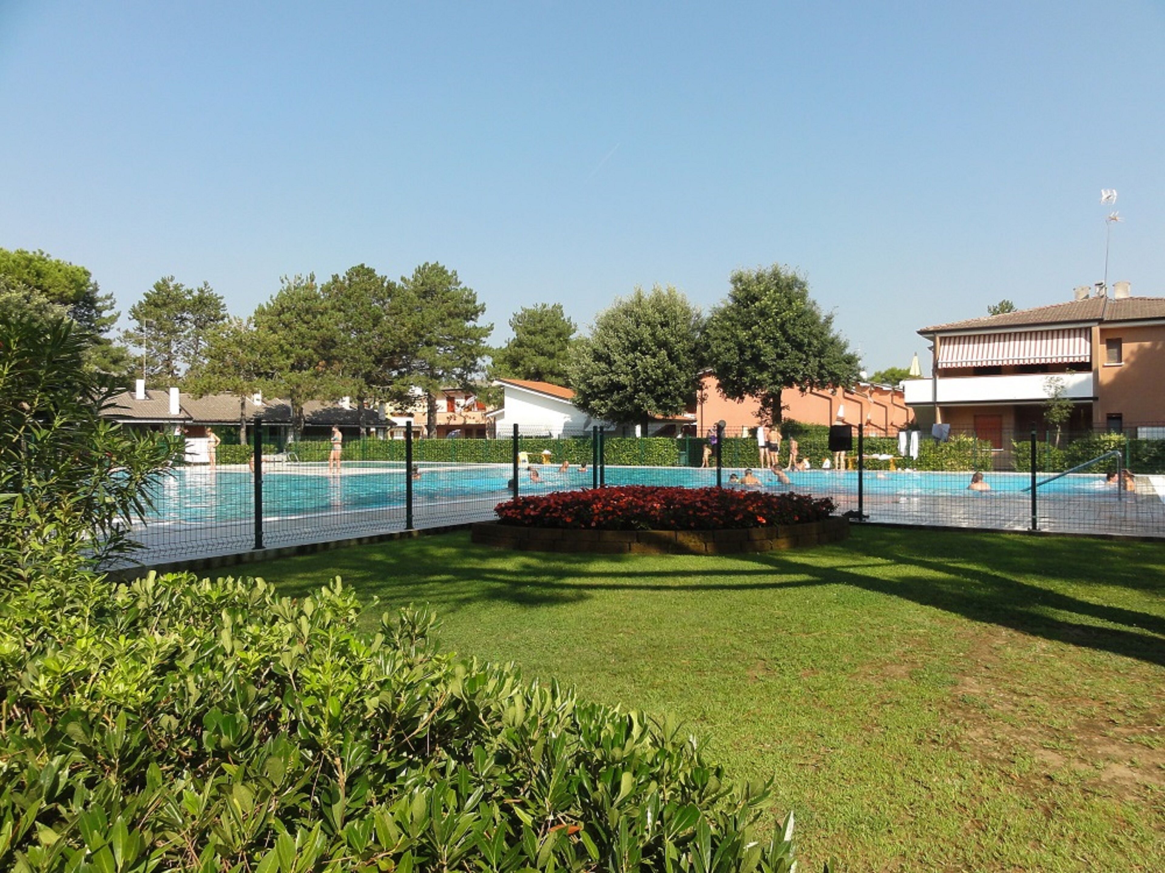 Piscina cercada por uma grade, com área gramada em primeiro plano e edifícios ao fundo.
