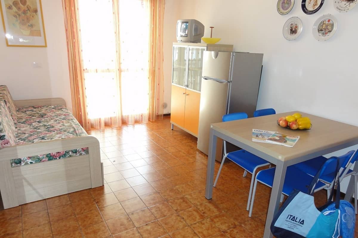 Quarto pequeno e bem iluminado, com cama, mesa e cadeiras, geladeira e janela com cortinas.