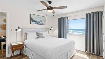 Quarto com cama grande, ventoinha no teto, mesa de cabeceira, luminária, pintura e janela com cortinas.
