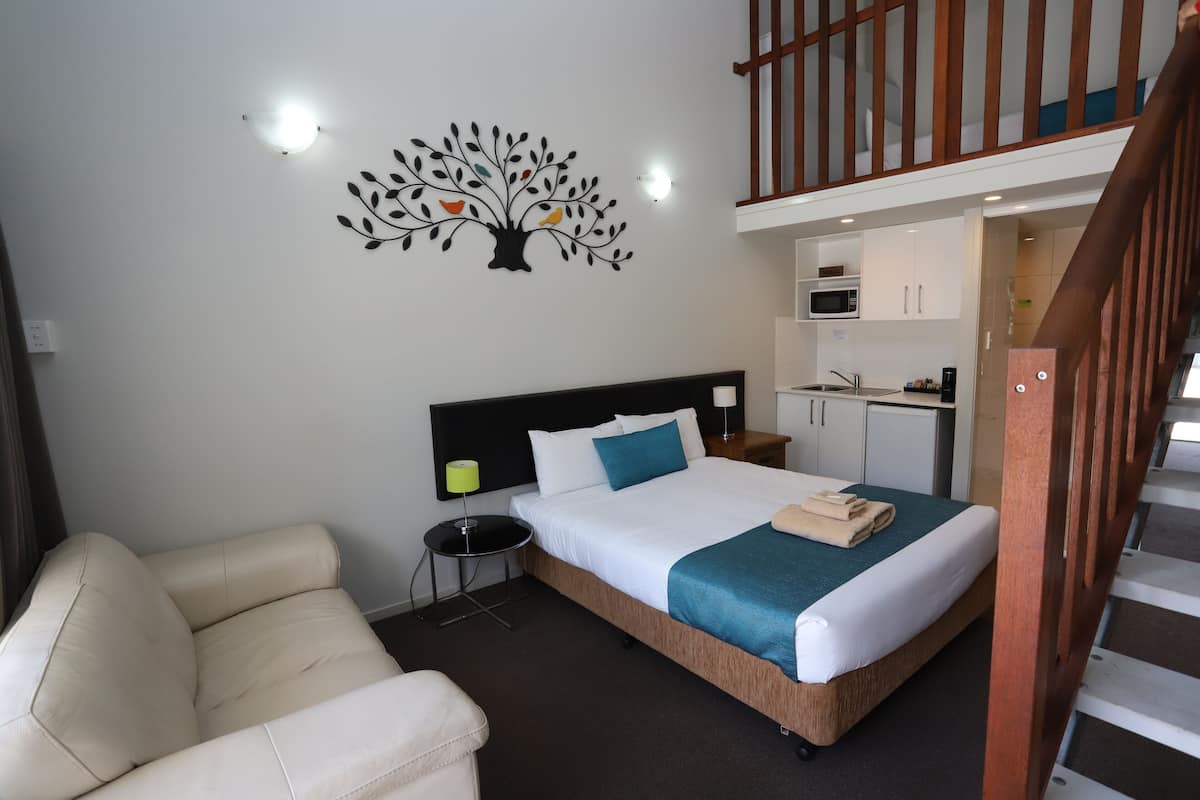 Quarto de hotel com cama, sofá, decoração de árvore na parede e área de cozinha compacta.