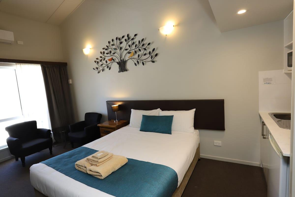 Quarto de hotel com uma cama grande, uma mesinha de cabeceira, um adorno de árvore na parede e uma janela com cortinas.