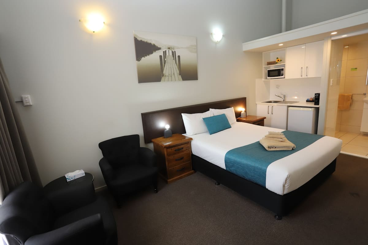 Quarto de hotel com cama, mesas de cabeceira, uma cadeira, escrivaninha, micro-ondas e uma pequena cozinha.