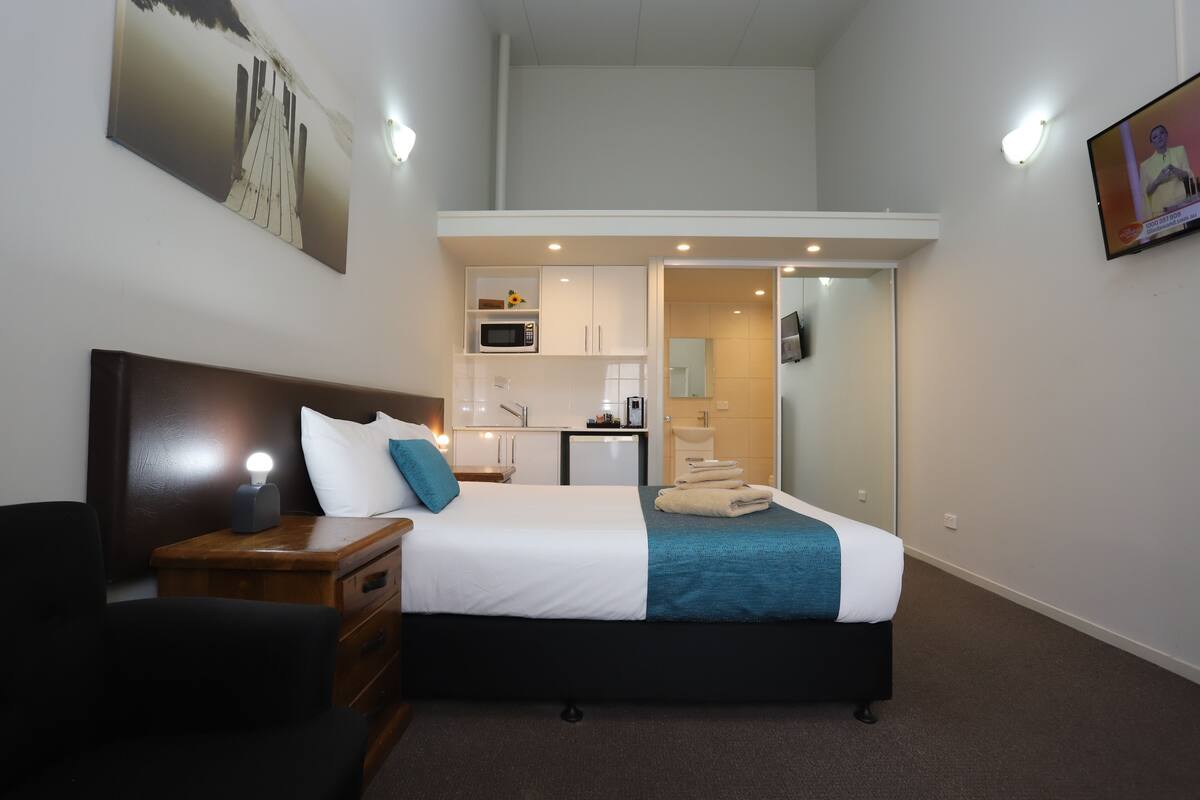 Quarto de hotel moderno com cama, mesa de cabeceira e vista para a cozinha.