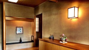 Interior tradicional japonês com lambris de madeira, um balcão de recepção, uma luminária na parede e uma cesta decorativa com flores.